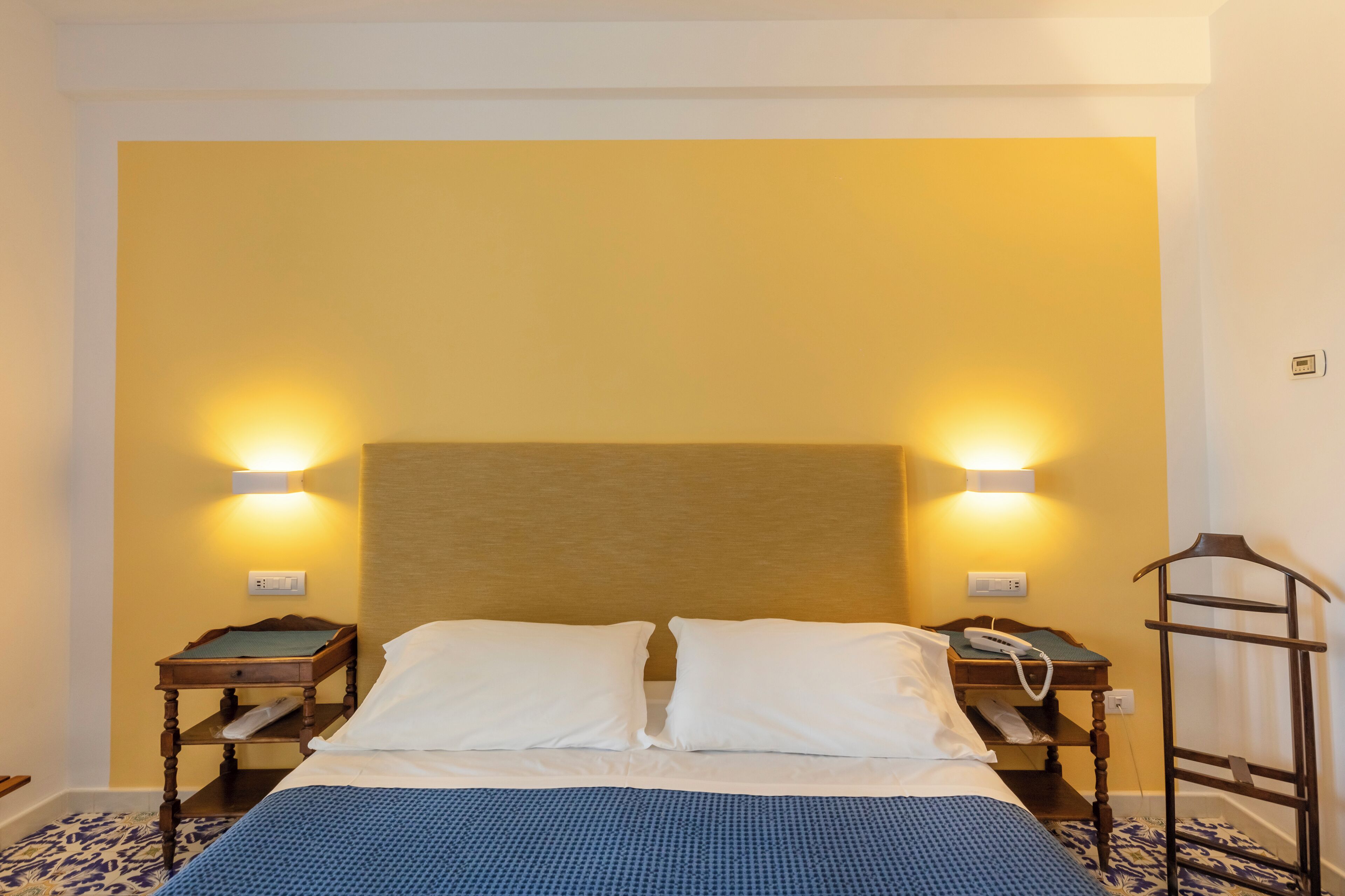 Quarto de hotel com cama, duas mesinhas de cabeceira, um criado-mudo, telefone fixado na parede e luminária na parede.