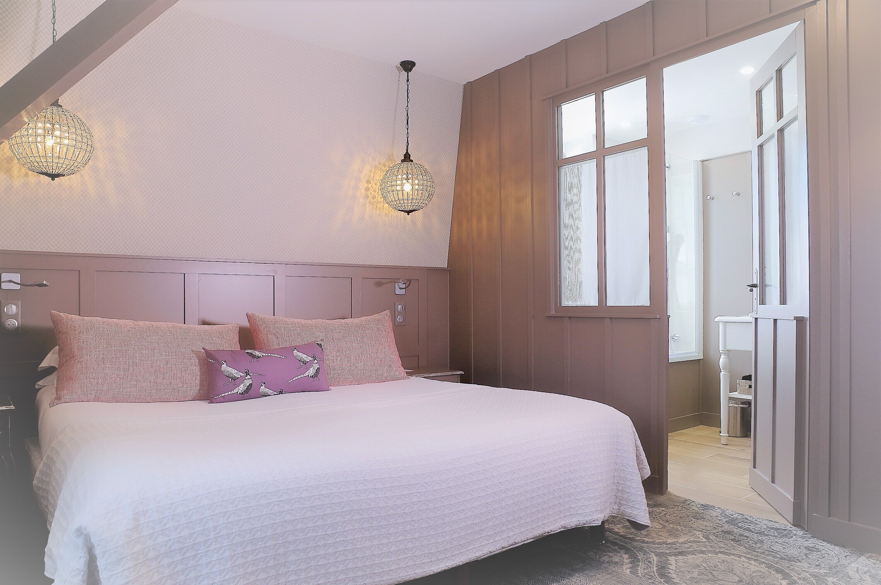 Quarto com uma cama grande, duas luminárias pendentes e vista para um banheiro através de uma porta.