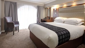 Quarto de hotel com uma cama grande, uma cadeira, uma escrivaninha com telefone e um espelho.