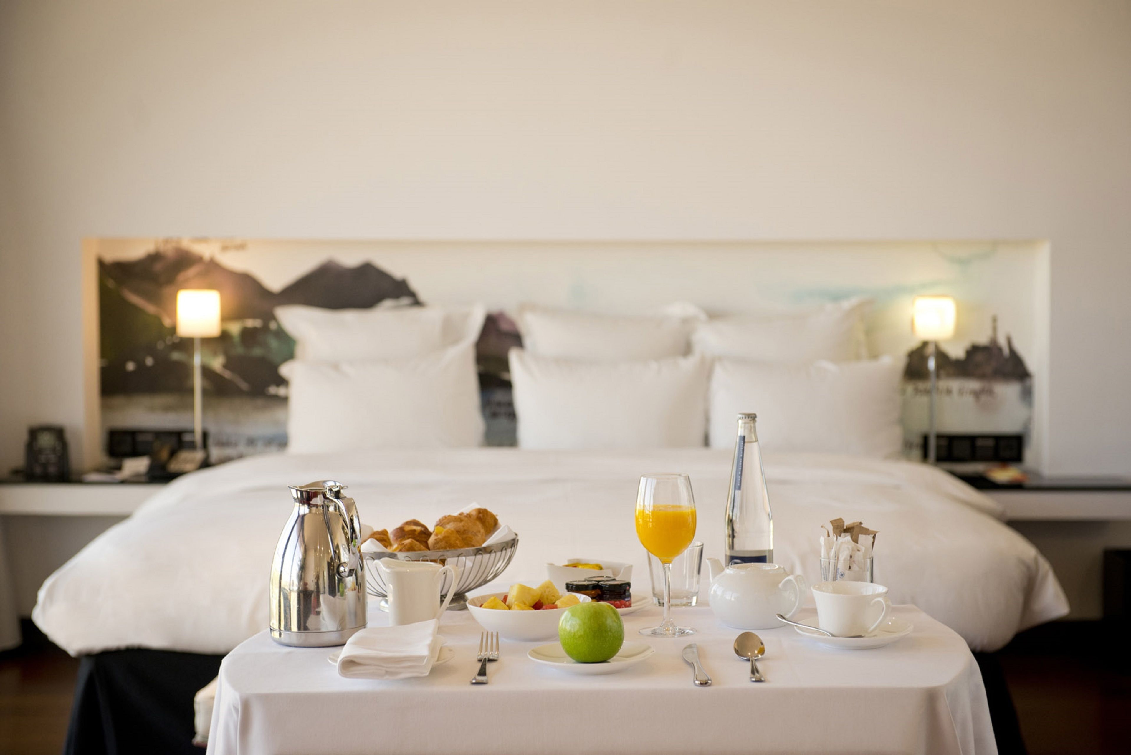 Mesa de café da manhã bem arrumada com croissants, frutas e suco de laranja em quarto de hotel com cama grande e vista para a cidade.