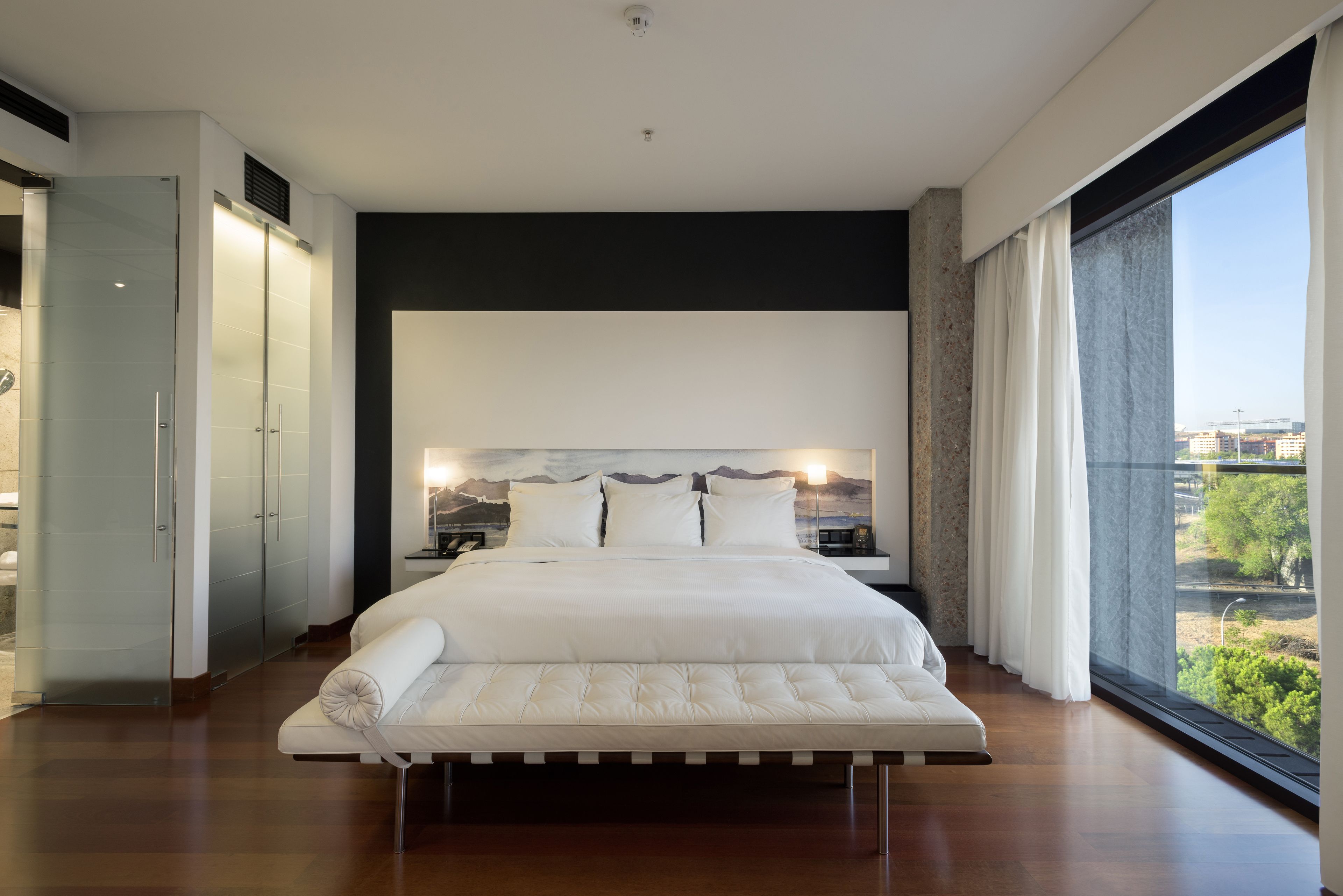 Quarto moderno com uma cama grande, uma mesa de cabeceira e vista da cidade através de amplas janelas.