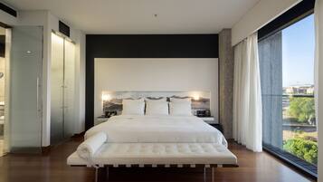 Quarto moderno com uma cama grande, uma mesa de cabeceira e vista da cidade através de amplas janelas.