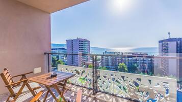 Varanda com mesa e cadeiras, guarda-corpo de vidro e vista para edifícios e água.