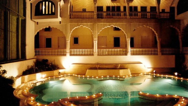 Piscina iluminada com um edifício com varandas e arcos.