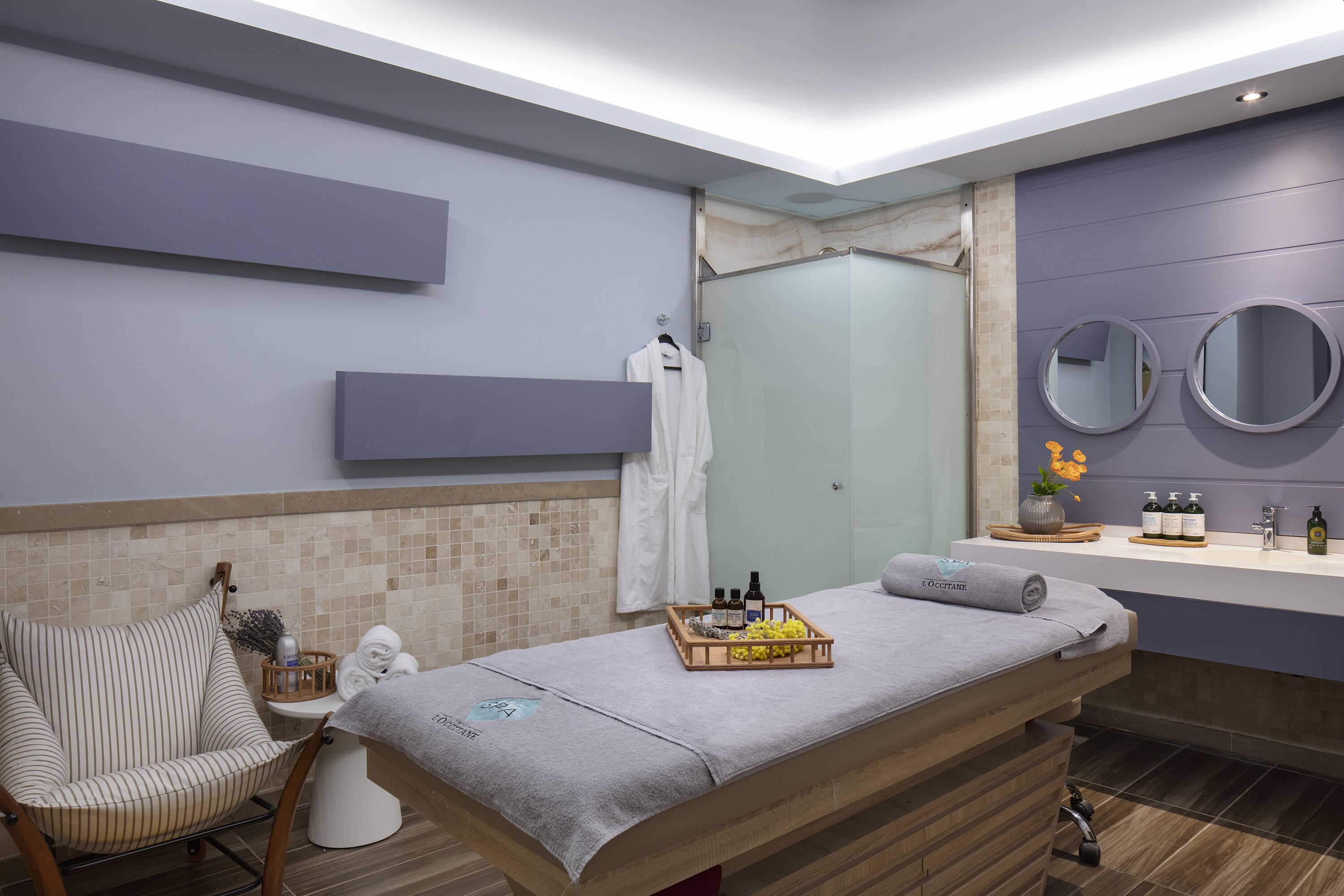 Uma sala de spa moderna com uma mesa de massagem, uma cadeira listrada, uma pequena mesa com garrafas e toalhas, e um lavatório com espelhos e artigos de higiene pessoal.