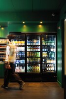 Uma pessoa caminhando por uma fileira de refrigeradores de bebidas em um quarto com paredes verdes e piso de madeira.