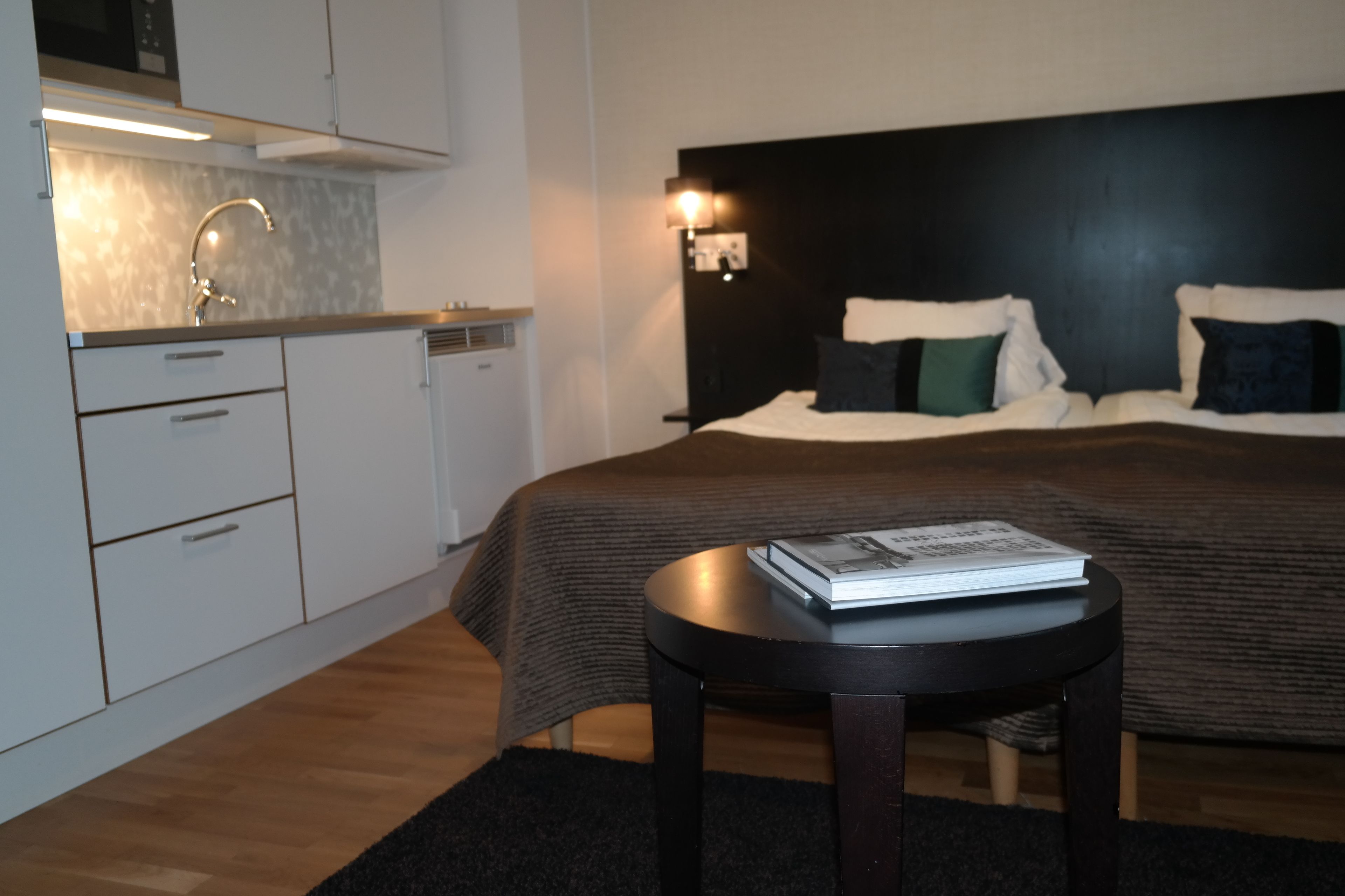 Quarto de hotel moderno com uma cama bem arrumada, uma pequena mesa redonda com um livro, área de cozinha com armários brancos e uma cabeceira escura.