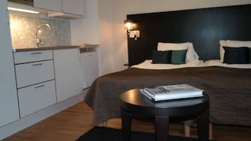 Quarto de hotel moderno com uma cama bem arrumada, uma pequena mesa redonda com um livro, área de cozinha com armários brancos e uma cabeceira escura.