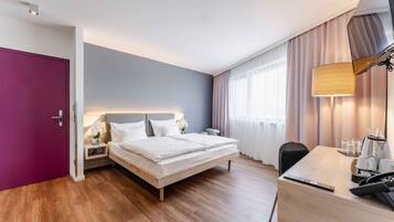 Quarto de hotel moderno com piso de madeira, cama com roupa de cama branca, escrivaninha com abajur e uma janela grande com cortinas.