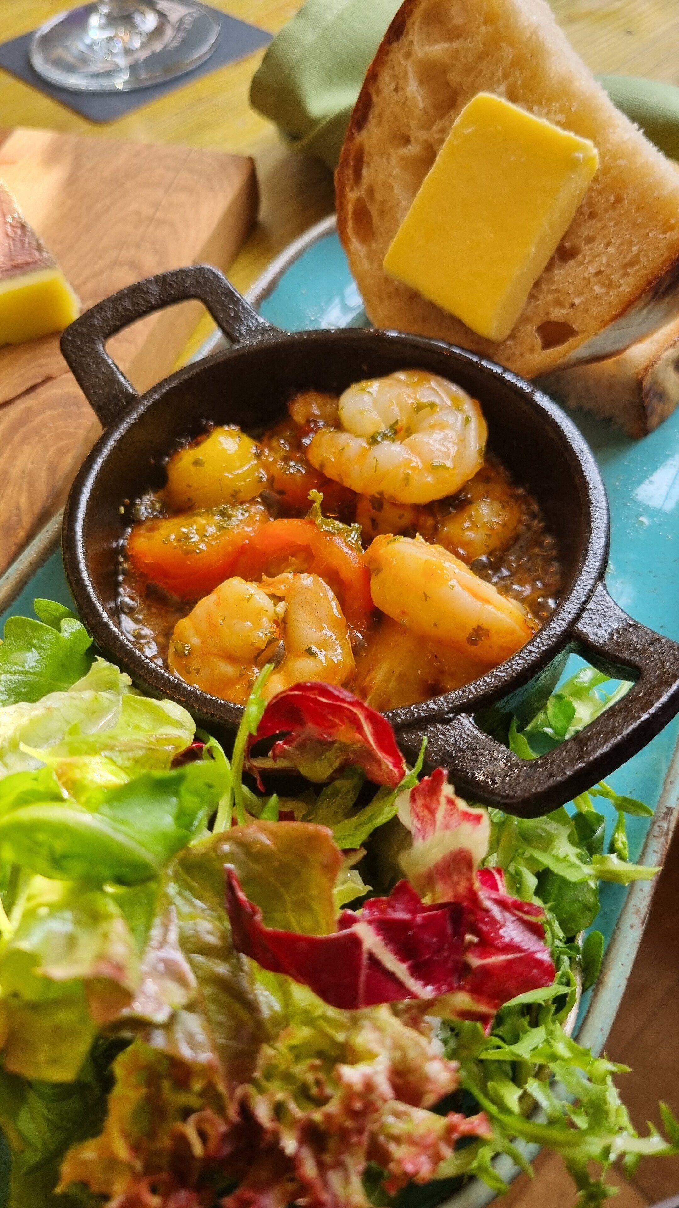 Uma refeição com camarões e legumes numa frigideira de ferro fundido, acompanhada de uma salada e fatias de pão.