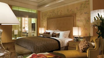 Quarto de hotel com uma cama grande, duas mesas de cabeceira, um espaço de estar com uma poltrona, uma mesa com uma revista e um vaso com flores.