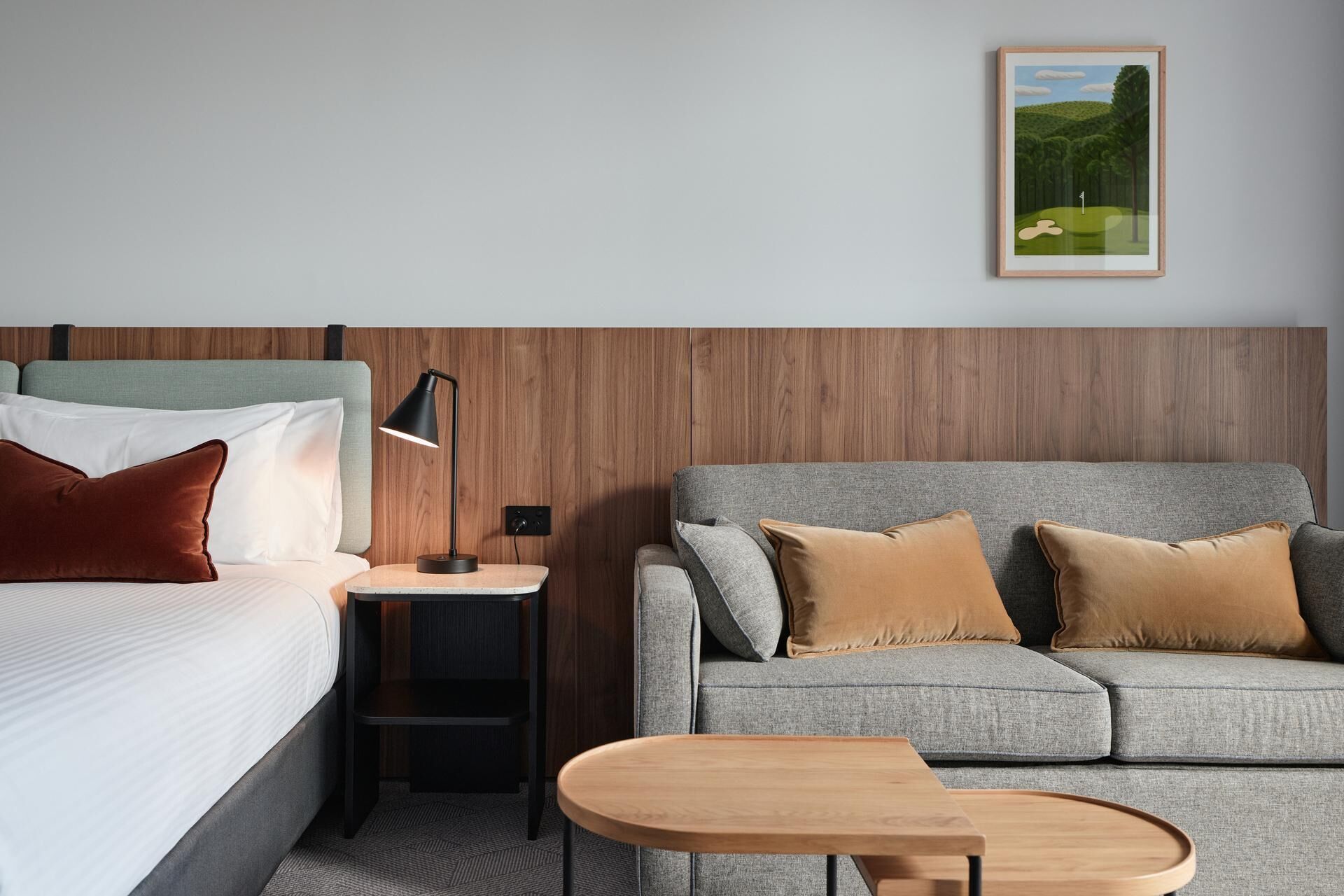 Quarto de hotel com cama, escrivaninha com luminária, sofá cinza com almofadas e um quadro de um campo de golfe.