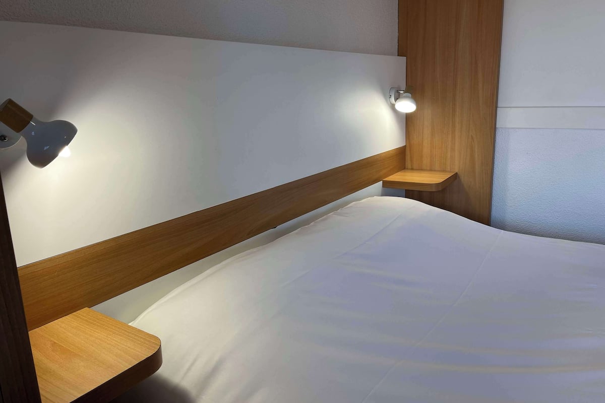 Uma cama bem arrumada com cabeceira de madeira, mesa de cabeceira e luminária fixada na parede.