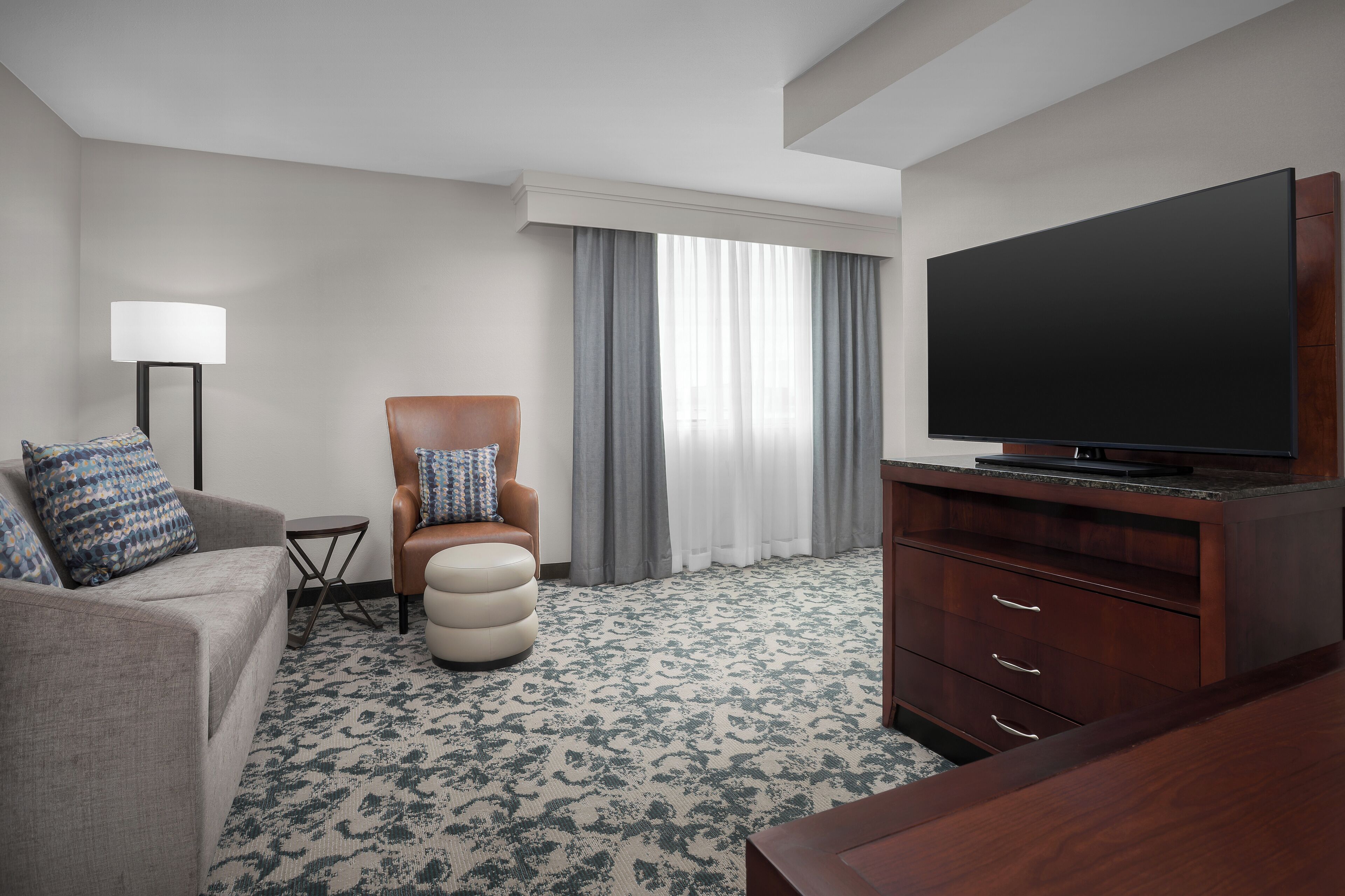 Quarto de hotel com sofá, poltrona, televisão e uma pequena mesa.