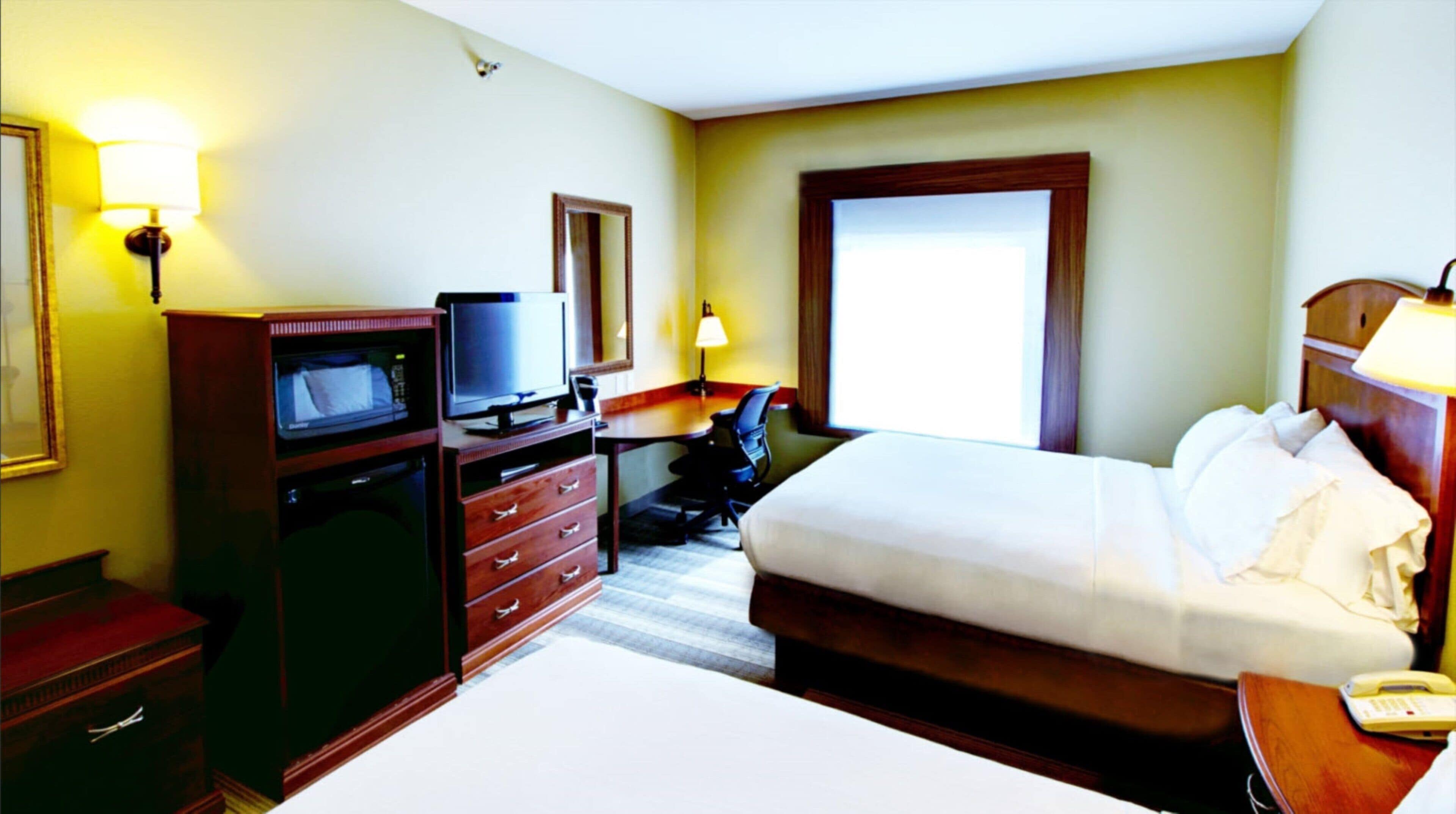 Quarto de hotel com cama, escrivaninha, computador, televisão e telefone.