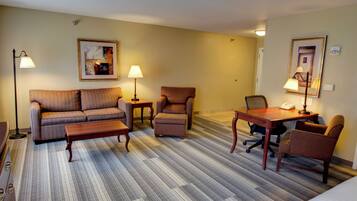 Quarto de hotel com sofá, poltrona, banqueta, uma mesinha e uma escrivaninha com cadeira. Há duas luminárias de chão e um quadro na parede.