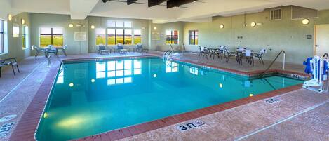 Uma piscina interior com água cristalina, rodeada por espreguiçadeiras e mesas, e grandes janelas com vista para o exterior.