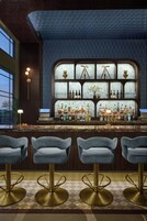 Uma área de bar com bancada de mármore, diversas garrafas de bebidas e três banquetas com base dourada.