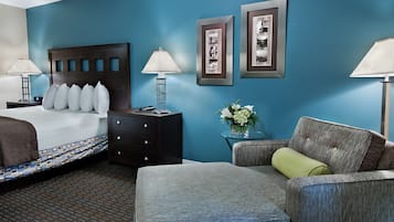 Quarto de hotel com cama, sofá, mesa de cabeceira, abajur e duas molduras na parede.