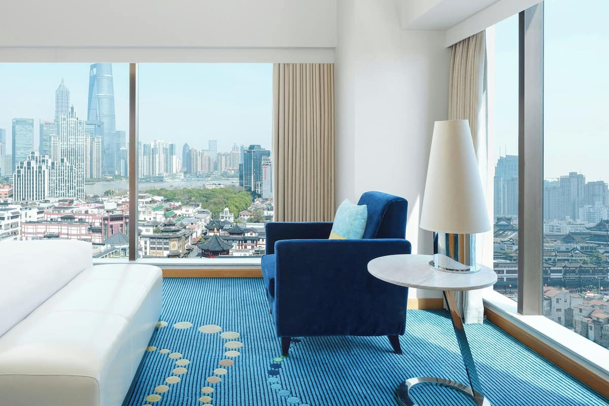 Quarto de hotel com uma grande janela com vista para a cidade, uma poltrona azul, um sofá branco e uma mesa pequena com abajur.