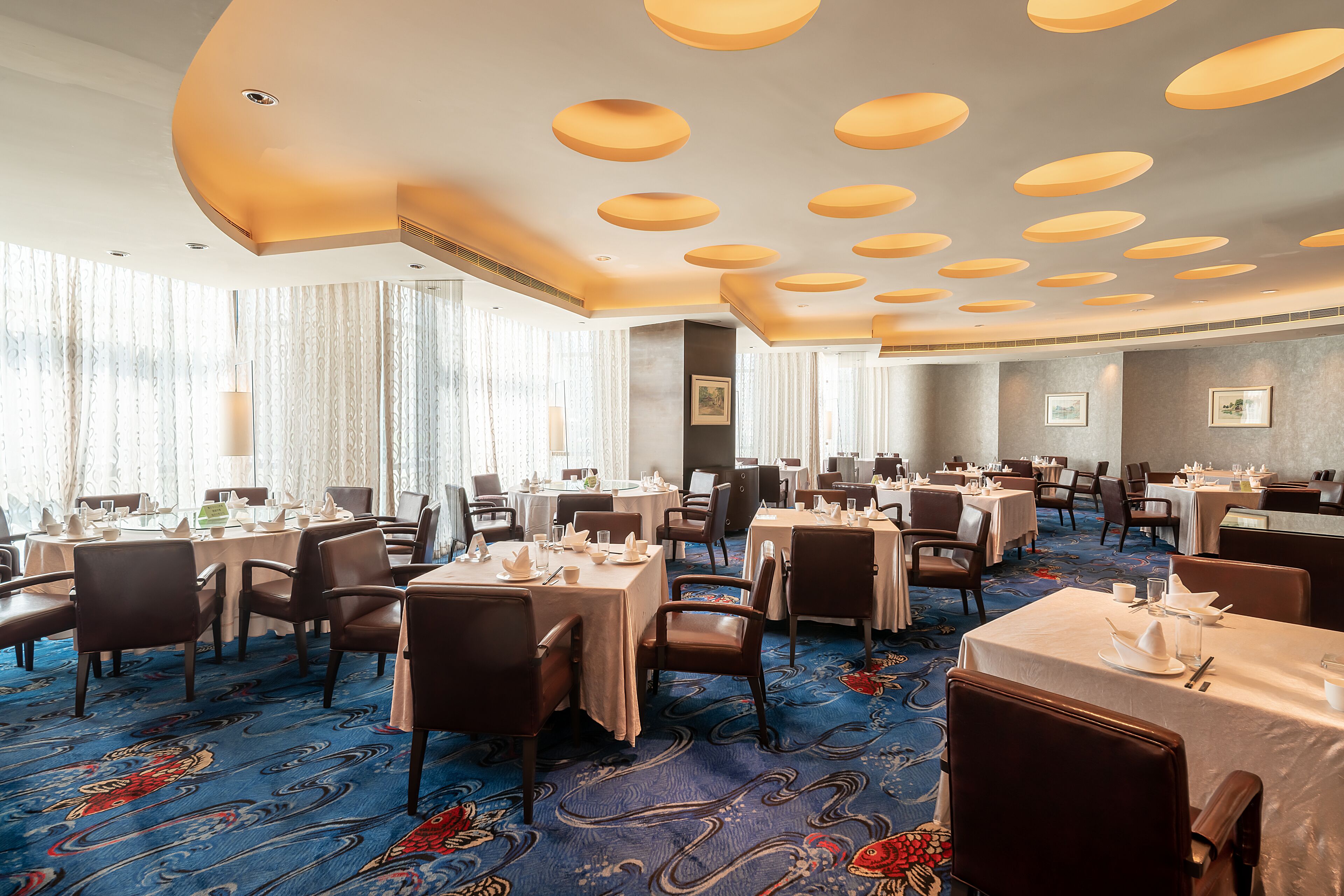 Espaço de refeições de hotel com mesas redondas, cadeiras e um tapete estampado.