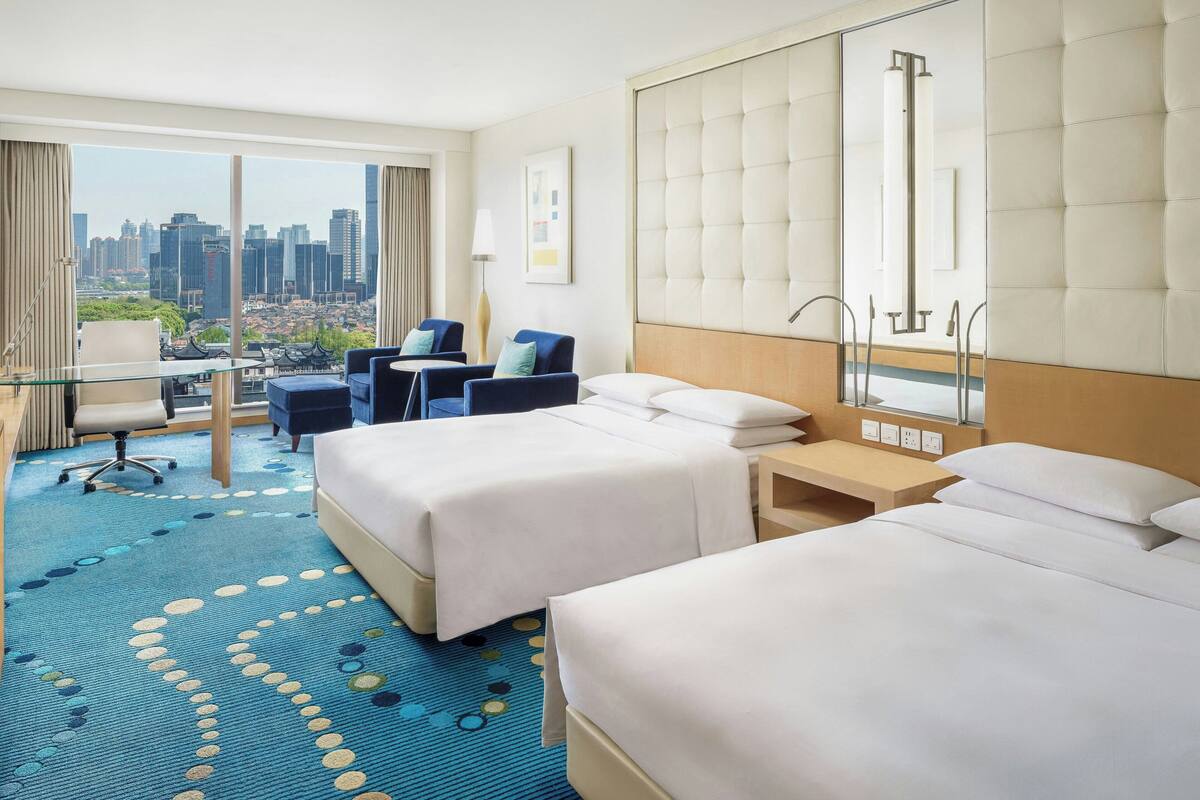 Quarto de hotel com duas camas, uma escrivaninha e uma cadeira. Tem uma janela grande com vista para a cidade.