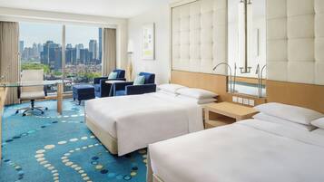 Quarto de hotel com duas camas, uma escrivaninha e uma cadeira. Tem uma janela grande com vista para a cidade.