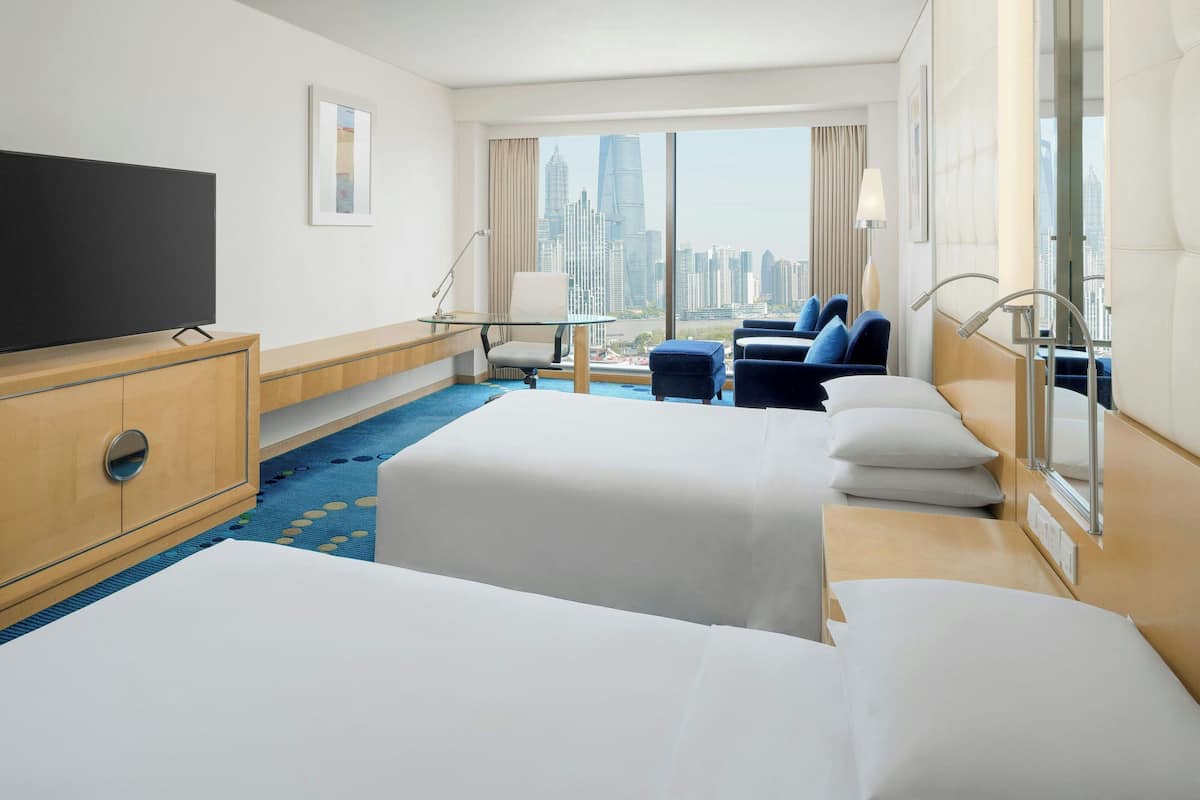 Quarto de hotel com duas camas, televisão, escrivaninha e vista para o horizonte urbano.