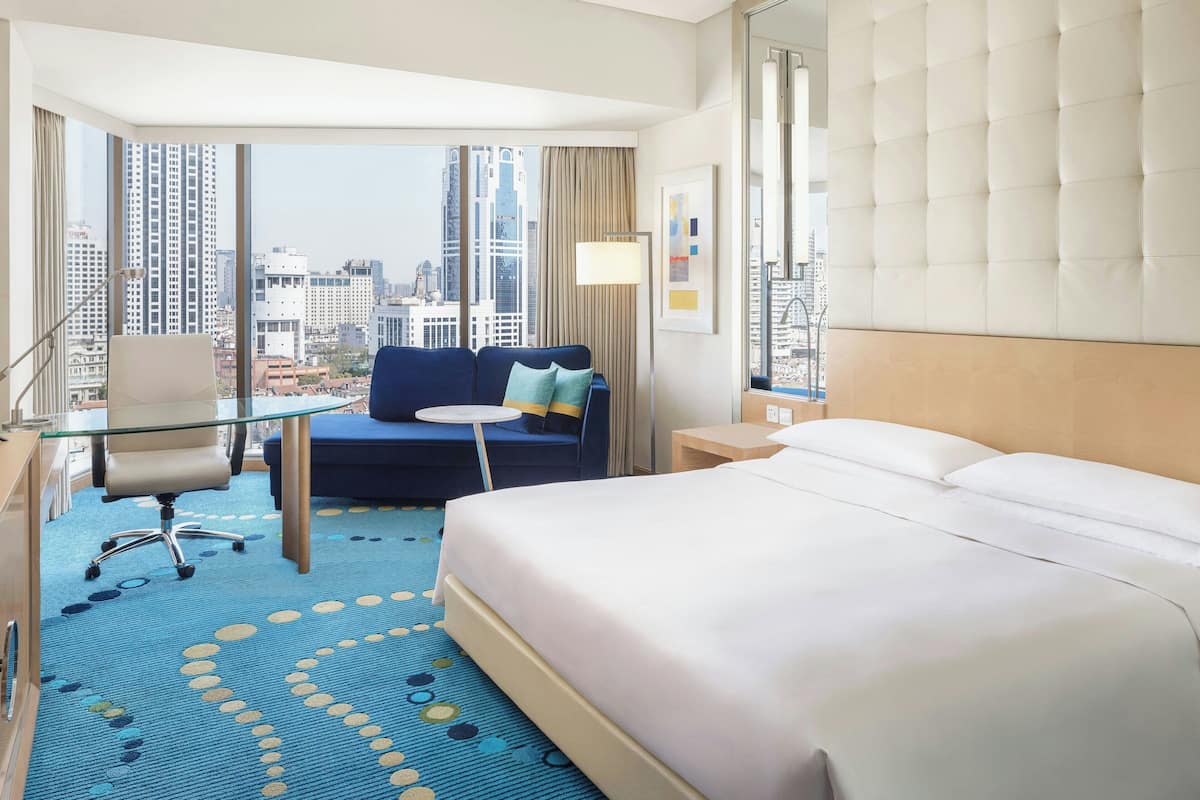 Quarto de hotel com uma cama grande, um sofá, uma escrivaninha e uma cadeira. Janelas amplas oferecem vista para a cidade.