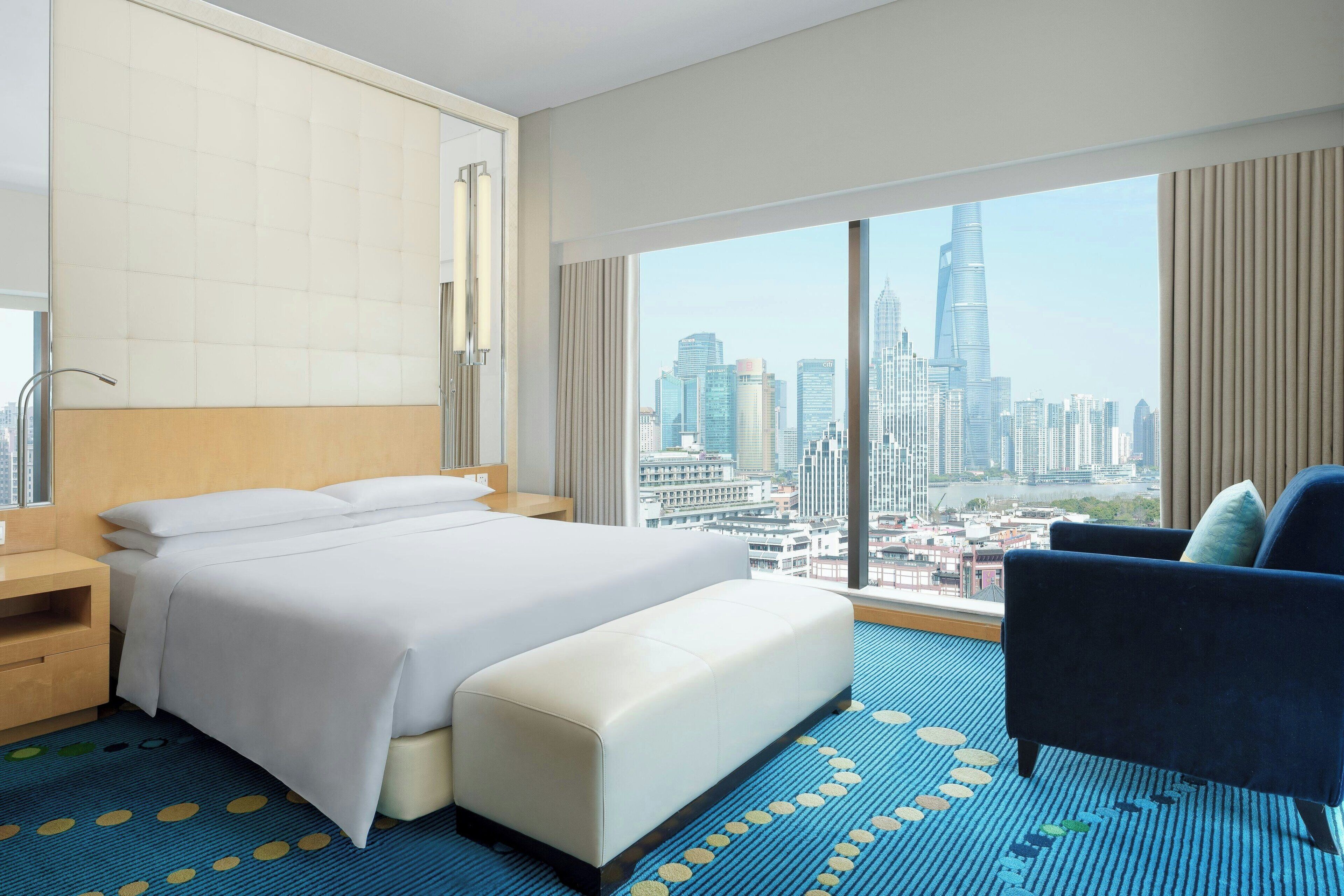 Quarto de hotel com uma cama grande, um banco, uma cadeira e vista para a cidade.