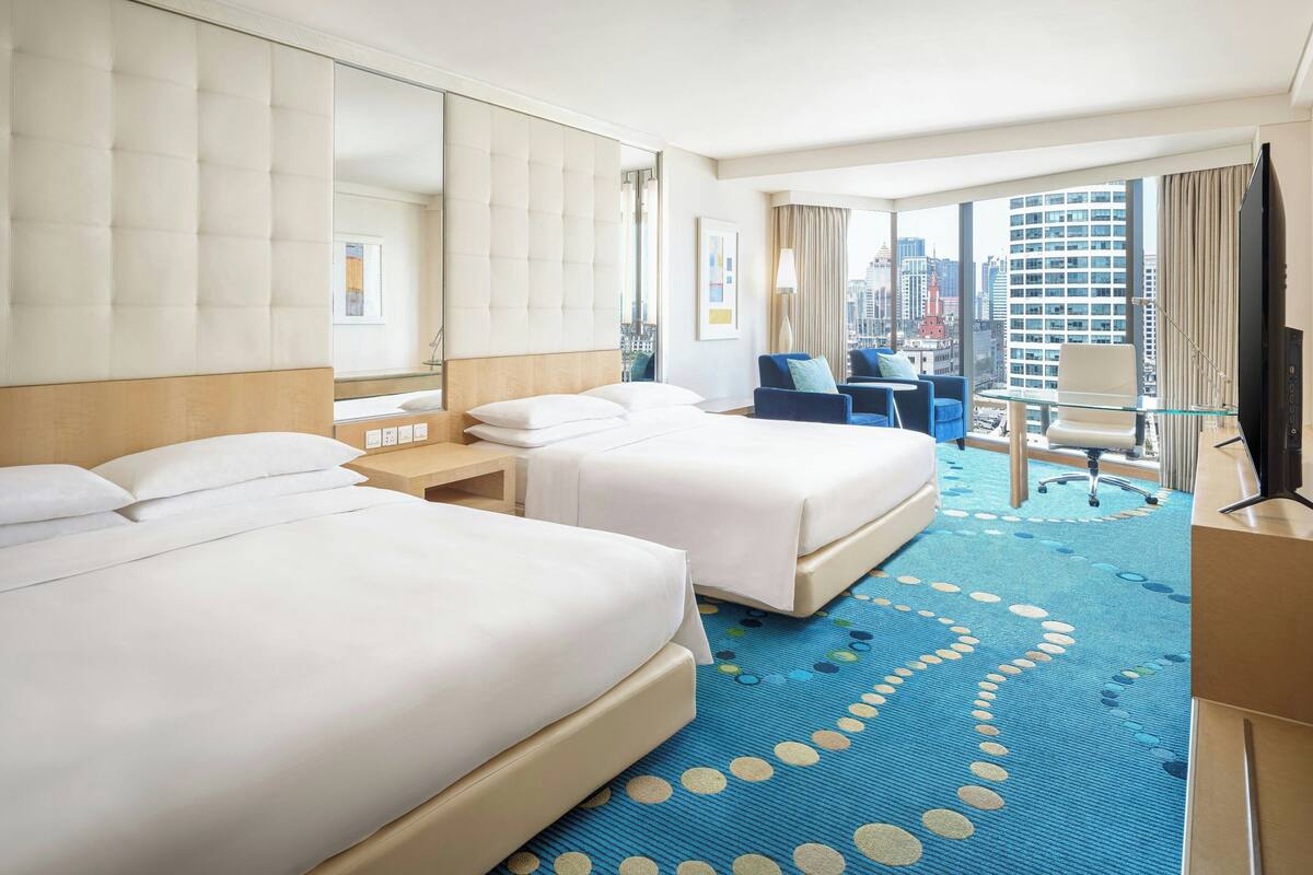 Quarto de hotel com duas camas, um espelho grande, uma escrivaninha, uma cadeira e uma televisão.