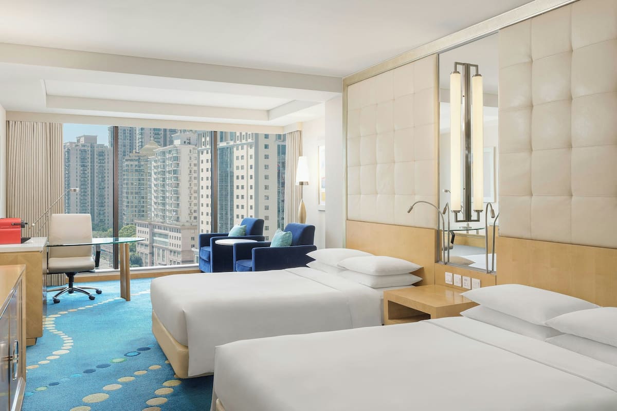 Quarto de hotel com duas camas, uma escrivaninha, uma cadeira e uma janela grande com vista para a cidade.