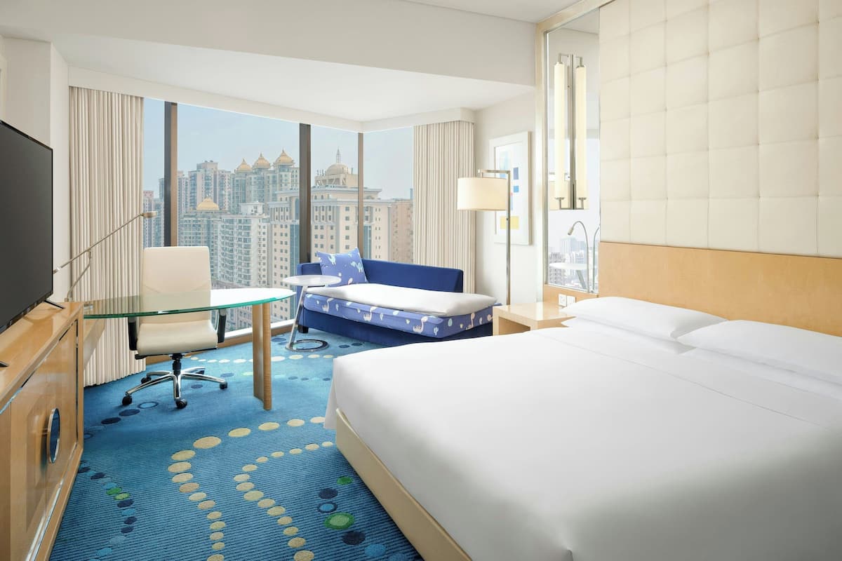 Quarto de hotel com uma cama grande, uma escrivaninha com cadeira, um sofá pequeno e vista para a cidade.