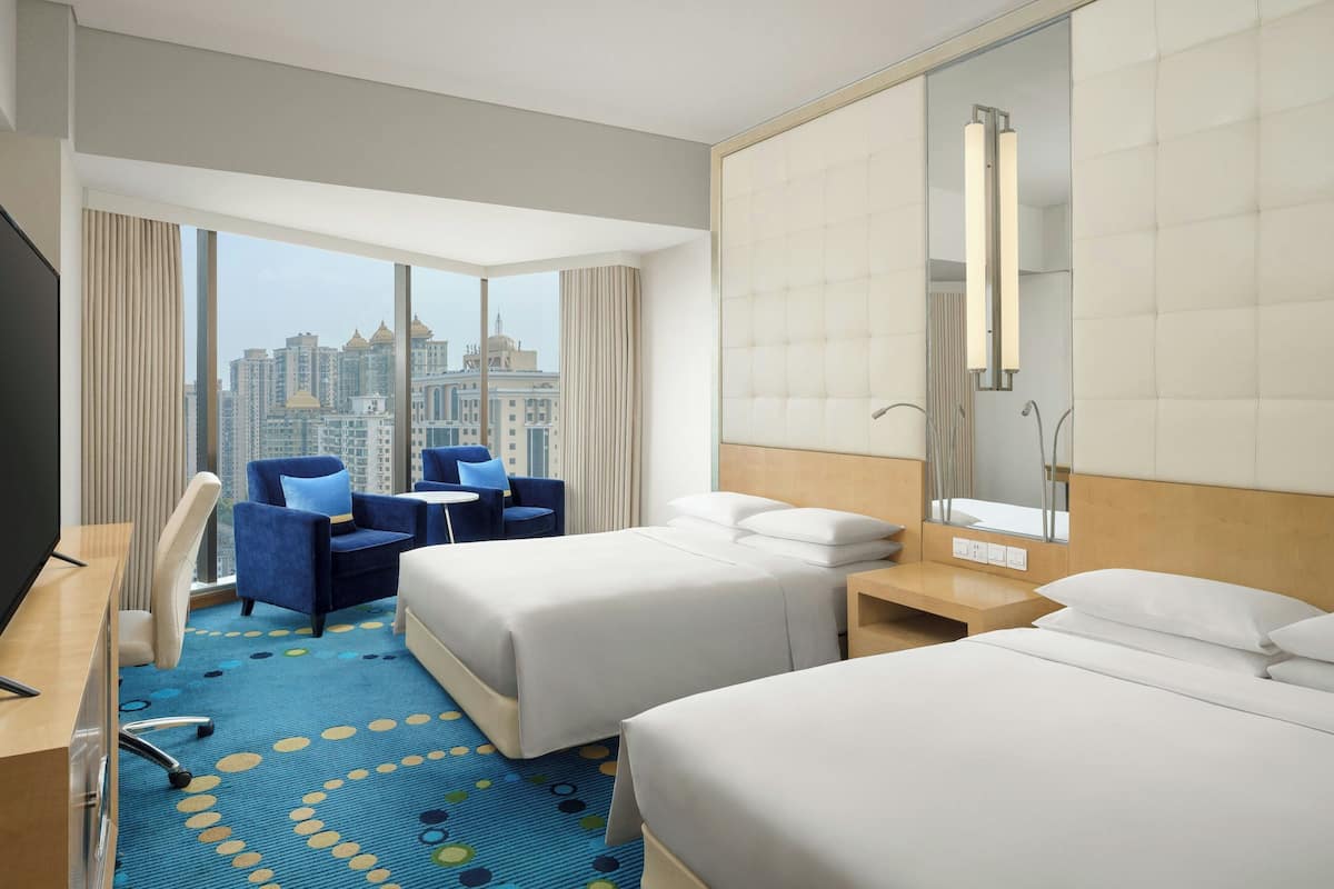 Quarto de hotel com duas camas, uma escrivaninha, uma cadeira e uma janela grande com vista para a cidade.