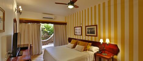Quarto de hotel com cama, mesa de cabeceira, abajur e televisão sobre uma escrivaninha.
