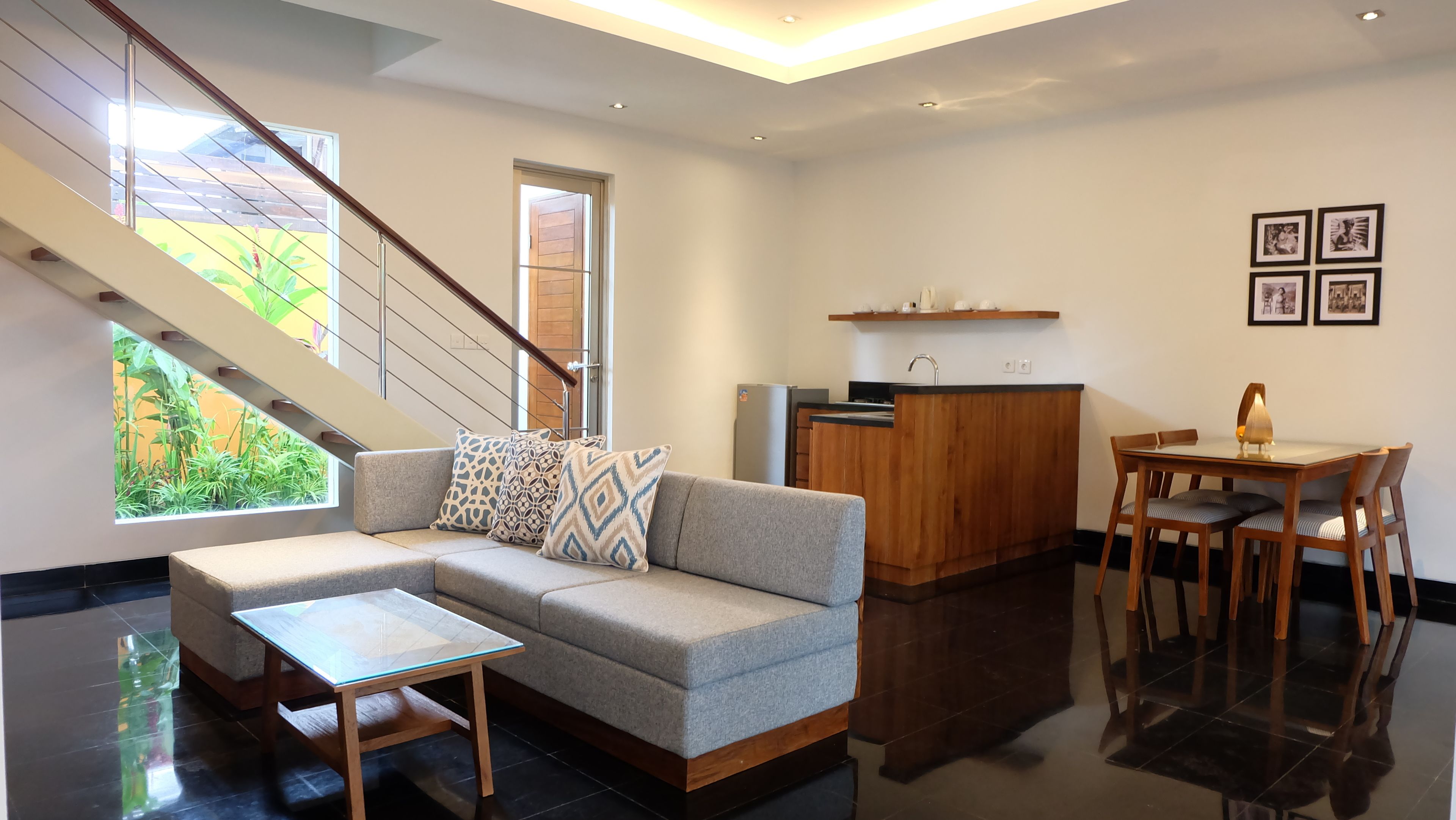 Uma sala de estar moderna com um sofá seccional cinza, uma mesa de centro de vidro, uma mesa de jantar de madeira com cadeiras, uma cozinha com bancada e pia de madeira, e uma escada com guarda-corpo de vidro que leva a uma varanda com plantas.