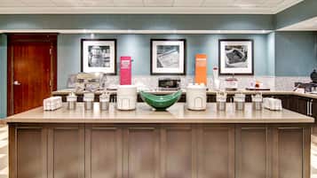 Uma área de recepção de hotel com um balcão amplo, quadros decorativos nas paredes e diversos serviços como máquinas de café e recipientes para temperos.