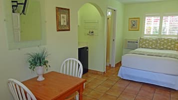 Quarto com cama, mesa de jantar com cadeiras, pequena geladeira e um vaso com planta.