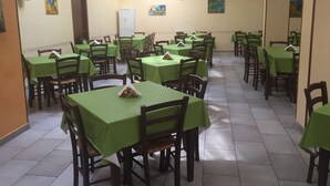 Uma sala de jantar com várias mesas cobertas por toalhas verdes, cadeiras de madeira e uma geladeira branca ao fundo.
