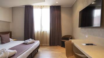 Quarto de hotel com cama, escrivaninha, cadeira, televisão e janela com cortinas.