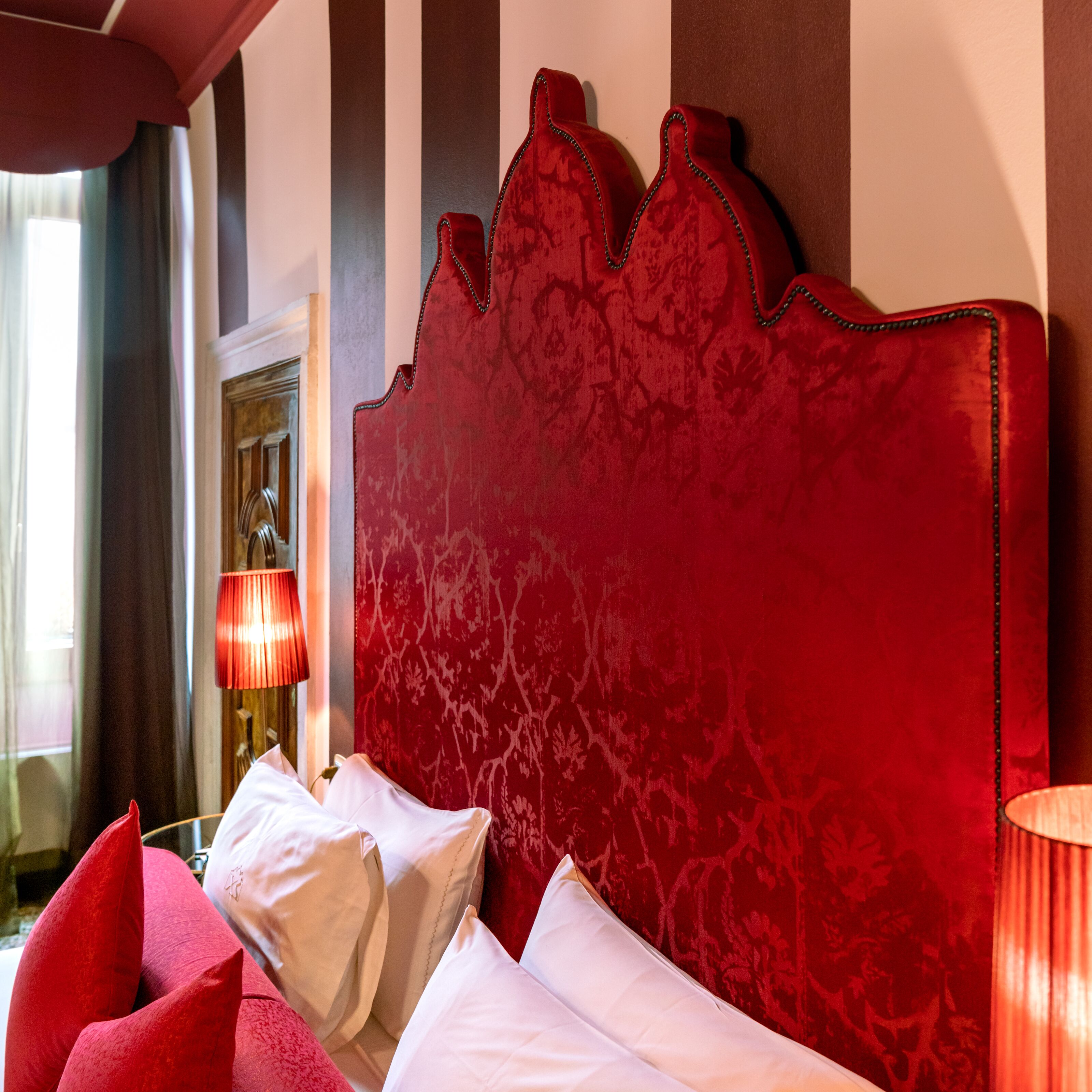 Quarto de hotel com uma cabeceira grande estofada em vermelho, roupa de cama branca e uma luminária vermelha na mesa lateral.