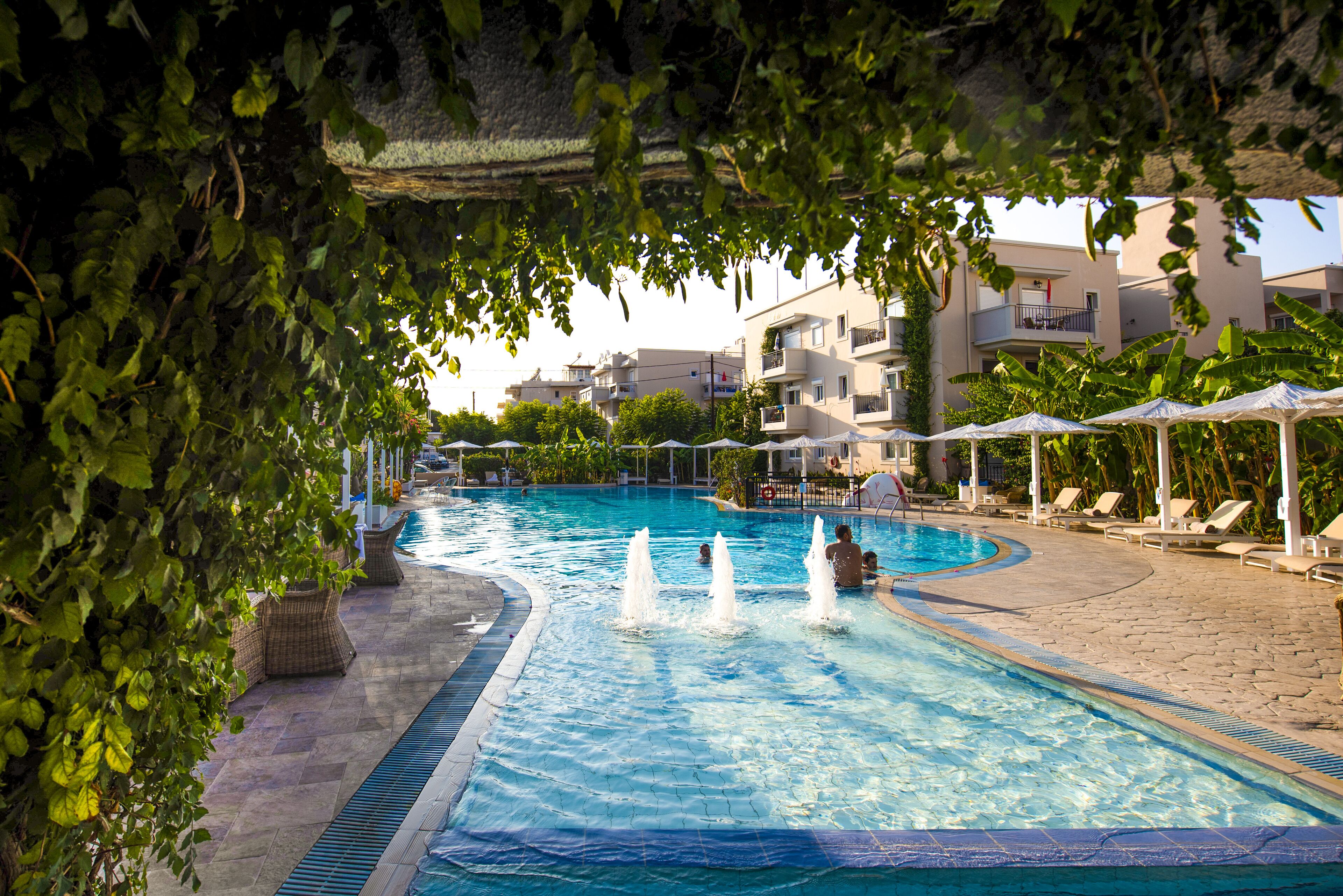 Piscina com fontes de água, rodeada por espreguiçadeiras e guarda-sóis, com edifícios ao fundo.