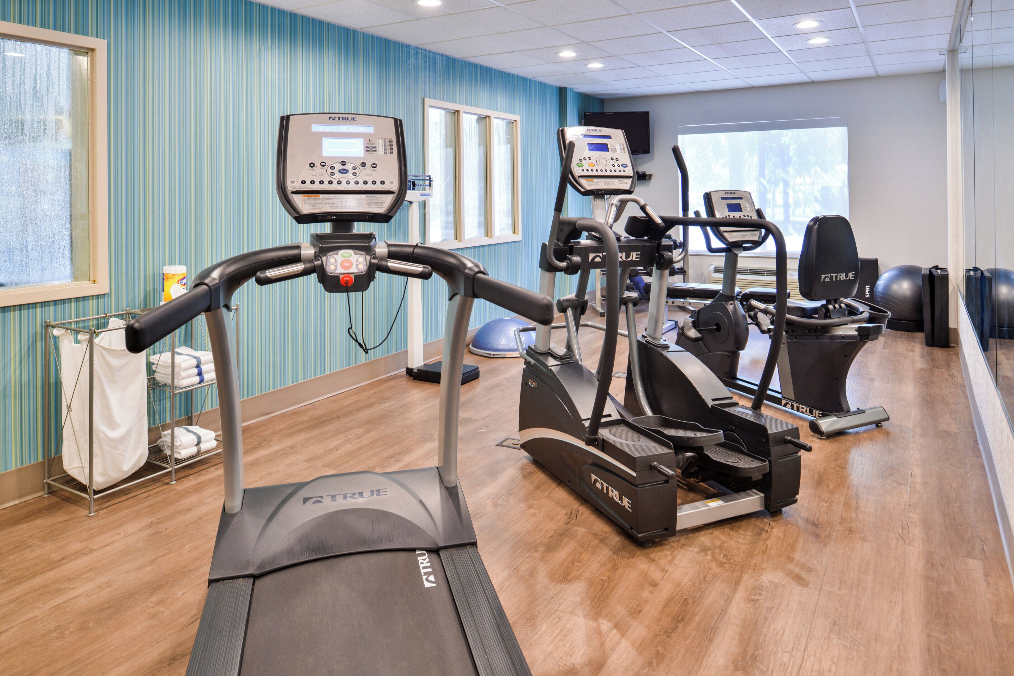 Uma academia com diversos equipamentos de exercícios, incluindo esteiras e elípticos, e um suporte com toalhas.
