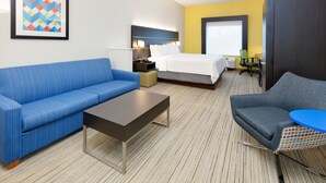 Quarto de hotel com sofá azul, poltrona cinza, uma mesinha, cama com roupa de cama branca, mesa de cabeceira com abajur e um quadro abstrato colorido na parede.