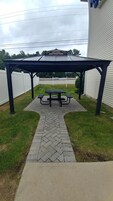 Uma área de piquenique coberta, com mesa e bancos, rodeada por um caminho pavimentado e gramado.