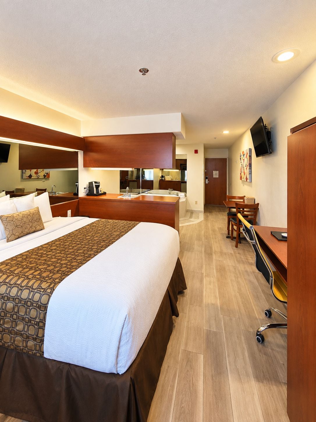 Quarto de hotel com cama grande, escrivaninha, cadeira, televisão e área de cozinha compacta.