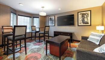 Quarto de hotel com área de estar, televisão de tela plana, janela grande e carpete estampado.