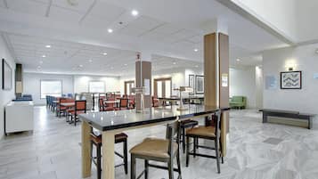Uma sala de jantar ampla, com várias mesas e cadeiras, teto alto com iluminação embutida e piso estampado.