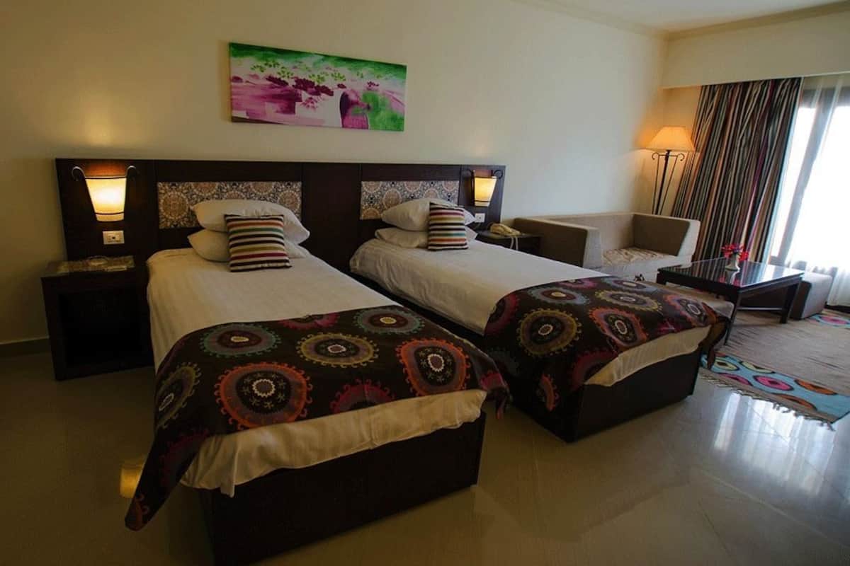 Quarto de hotel com duas camas, uma cadeira, uma mesinha de cabeceira e um abajur.