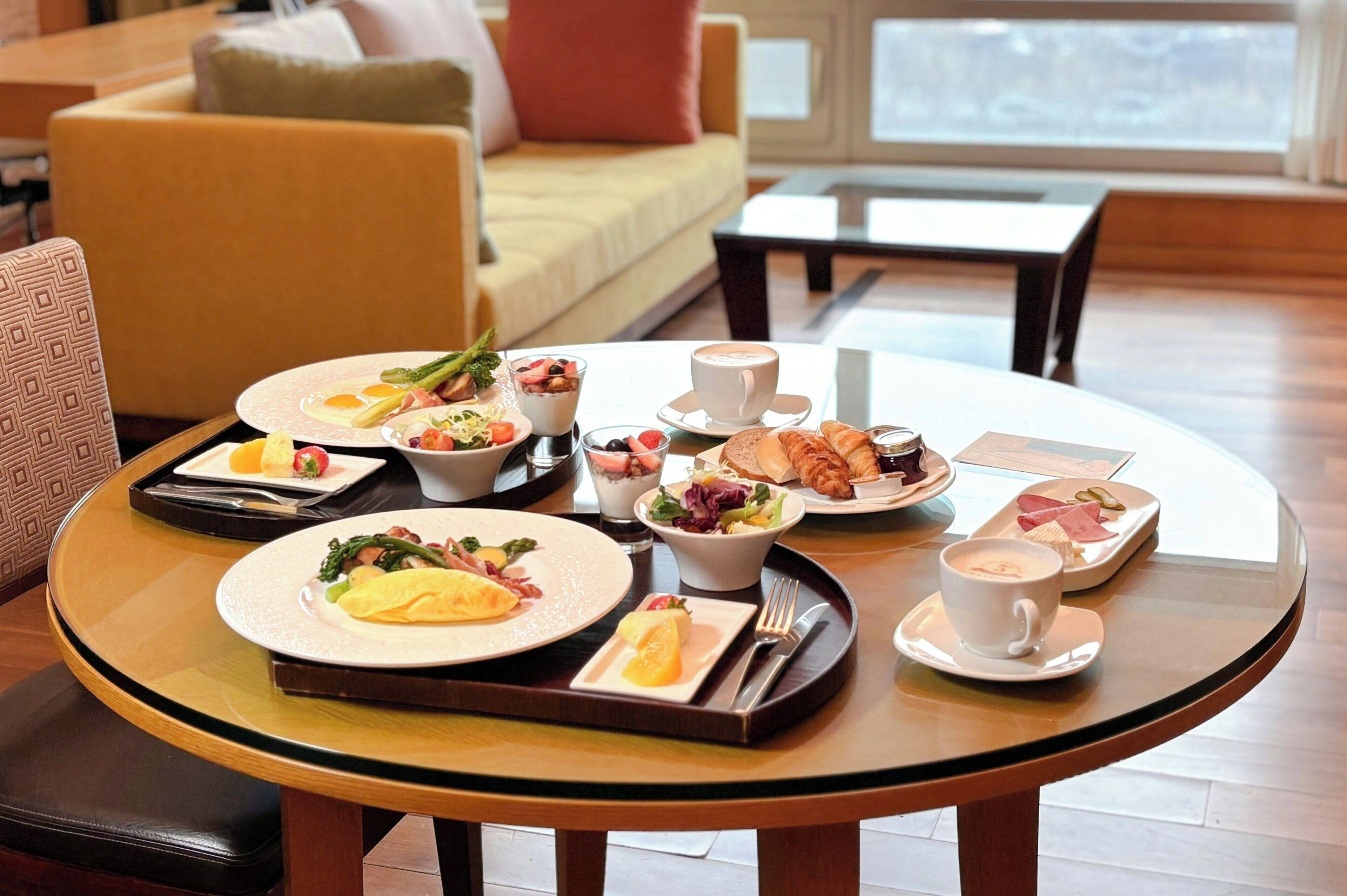 Disposição de café da manhã com diversos pratos, incluindo ovos, frutas e doces, sobre uma mesa redonda em um quarto de hotel moderno.